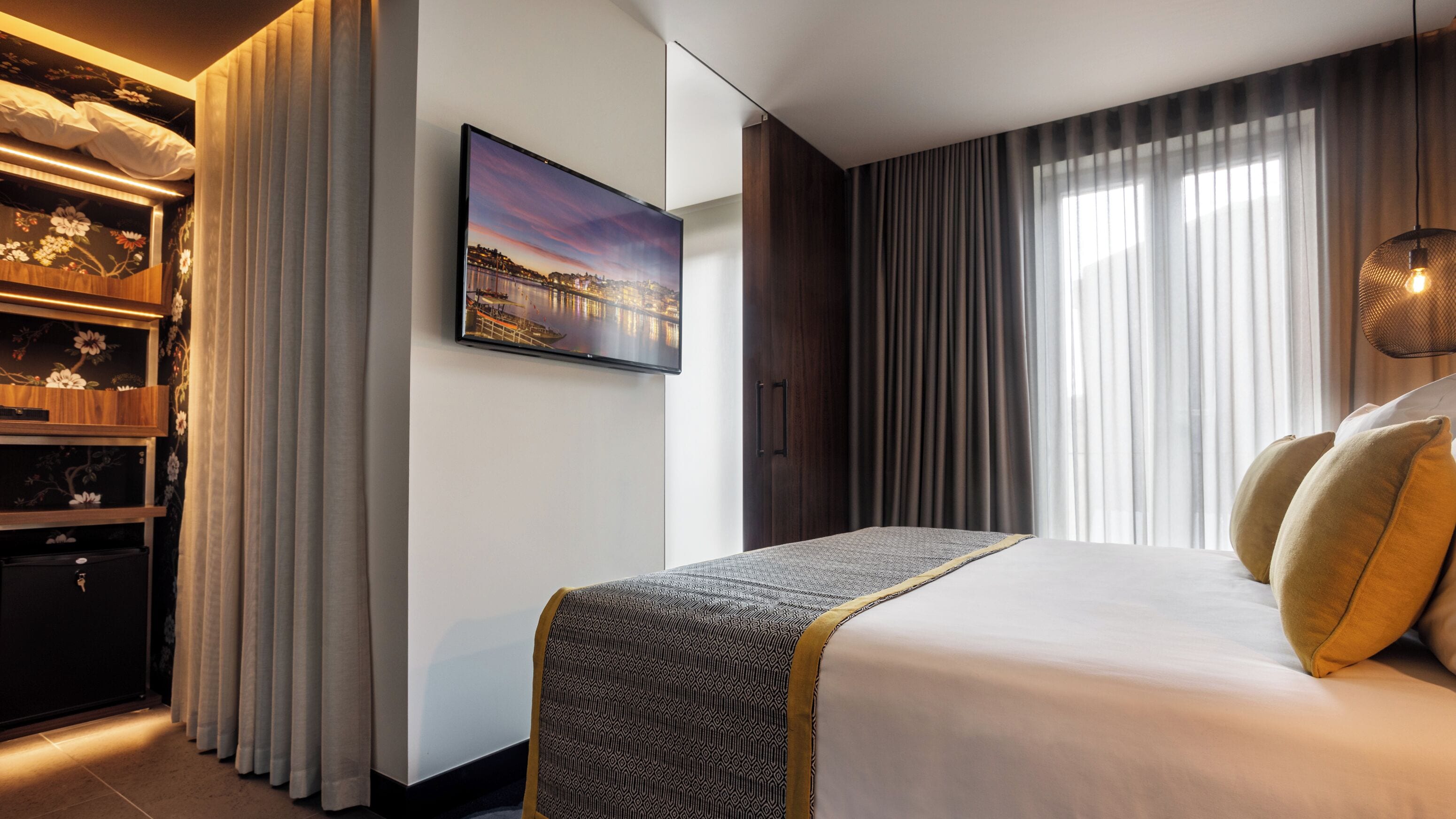 Quarto de hotel moderno com uma cama grande, televisão de tela plana na parede, guarda-roupa e vista para o exterior pela janela.