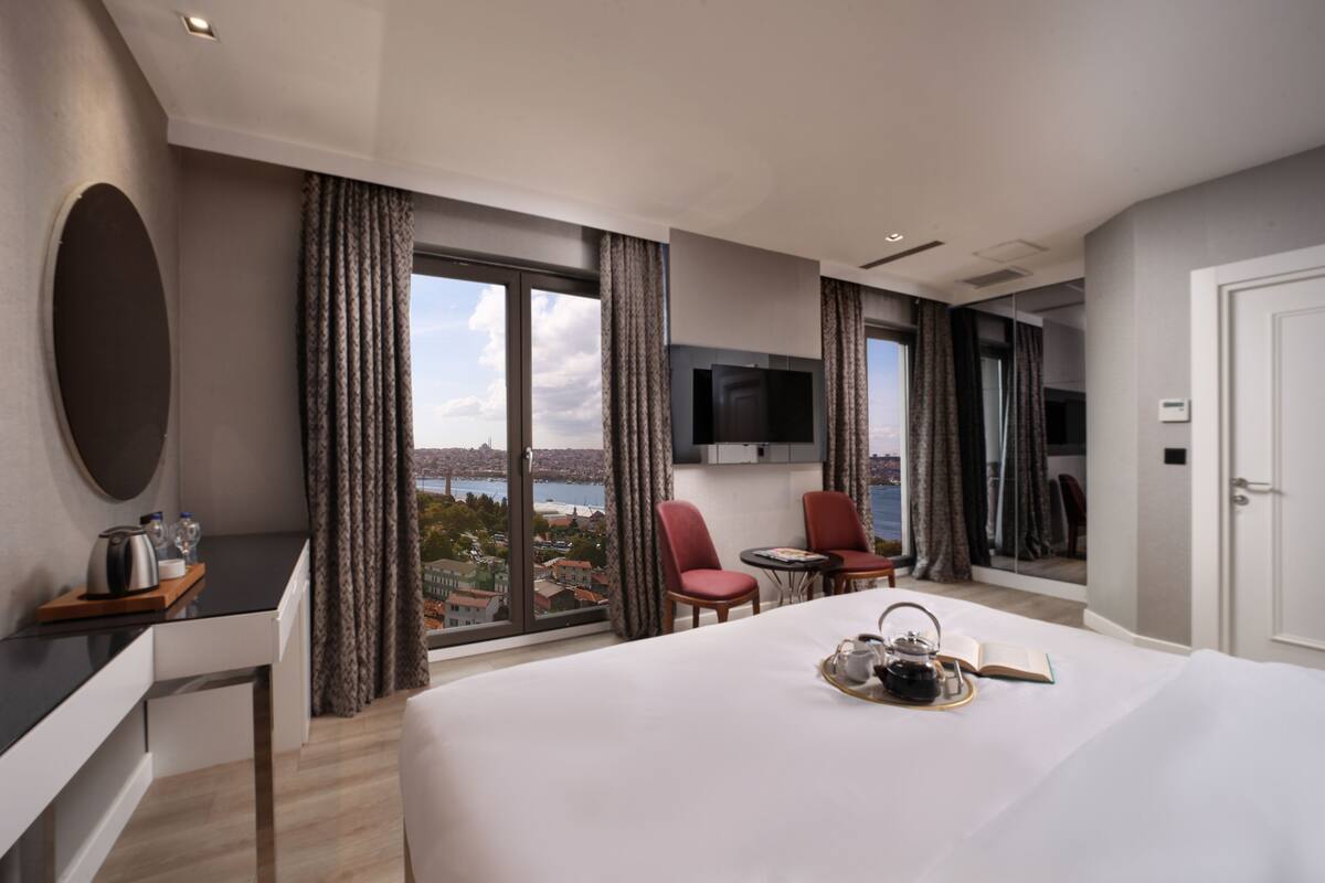 Quarto de hotel com uma cama grande, televisão fixada na parede, escrivaninha com chaleira e chávenas, duas cadeiras e vista para a cidade e para o rio.