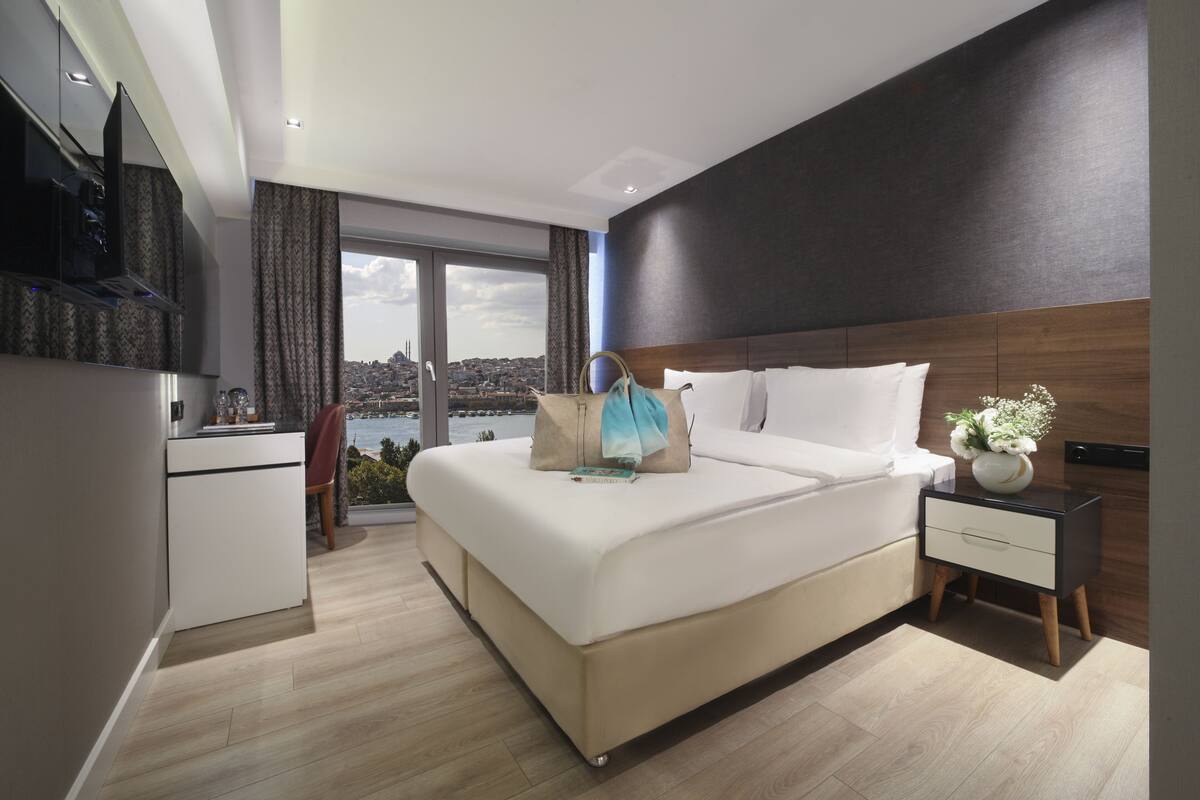 Quarto de hotel moderno com uma cama grande, mesa de cabeceira, escrivaninha e vista para a paisagem urbana através de uma janela ampla.