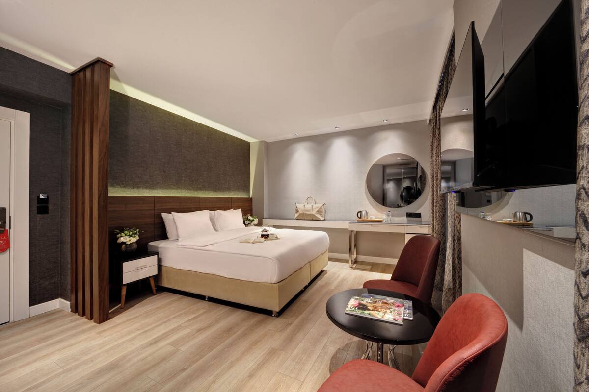 Quarto de hotel moderno com uma cama grande, uma escrivaninha com cadeira, uma TV e uma mesinha de centro com uma revista.