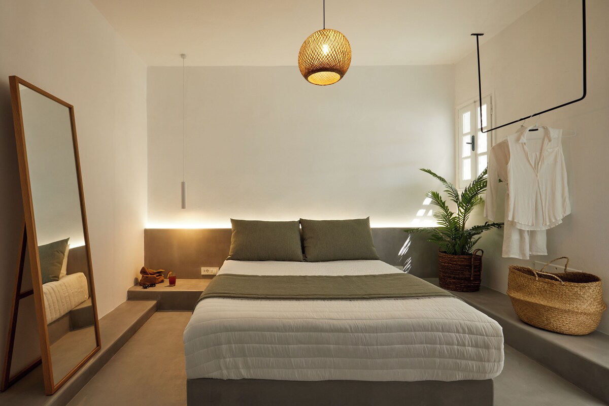 Quarto moderno com cama, mesa de cabeceira, espelho, luminária pendente, janela e planta em vaso.