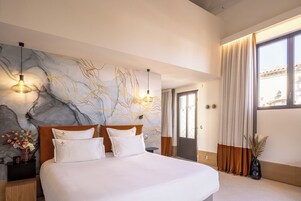 Quarto de hotel moderno com cama grande, parede de mármore com linhas douradas, janela com cortinas e mesa de cabeceira com um vaso de flores.