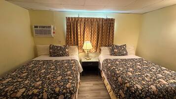Quarto de hotel com duas camas, uma mesa de cabeceira com abajur, cortinas e um aparelho de ar condicionado na parede.
