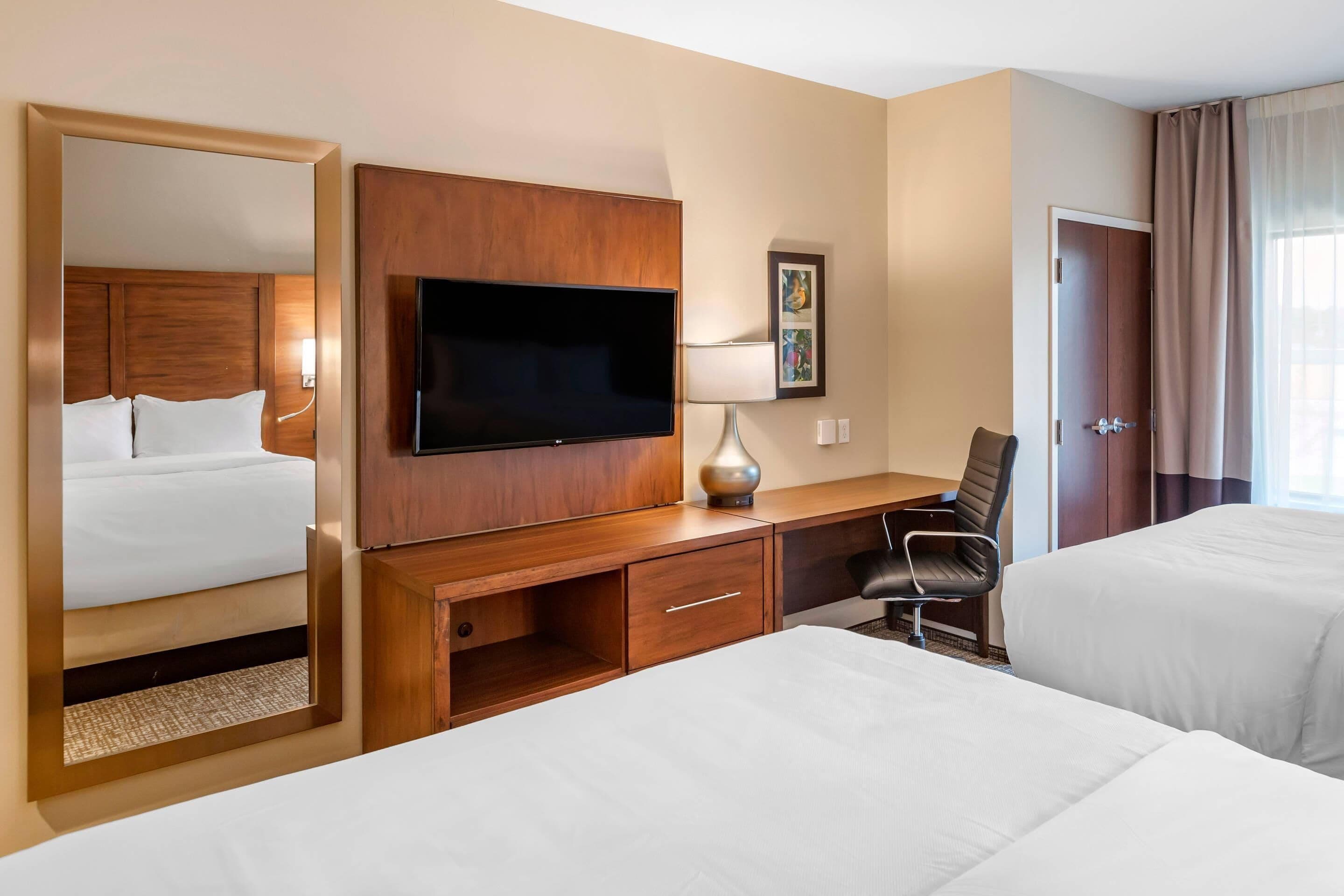 Quarto de hotel com cama, escrivaninha, televisão, cadeira e porta.