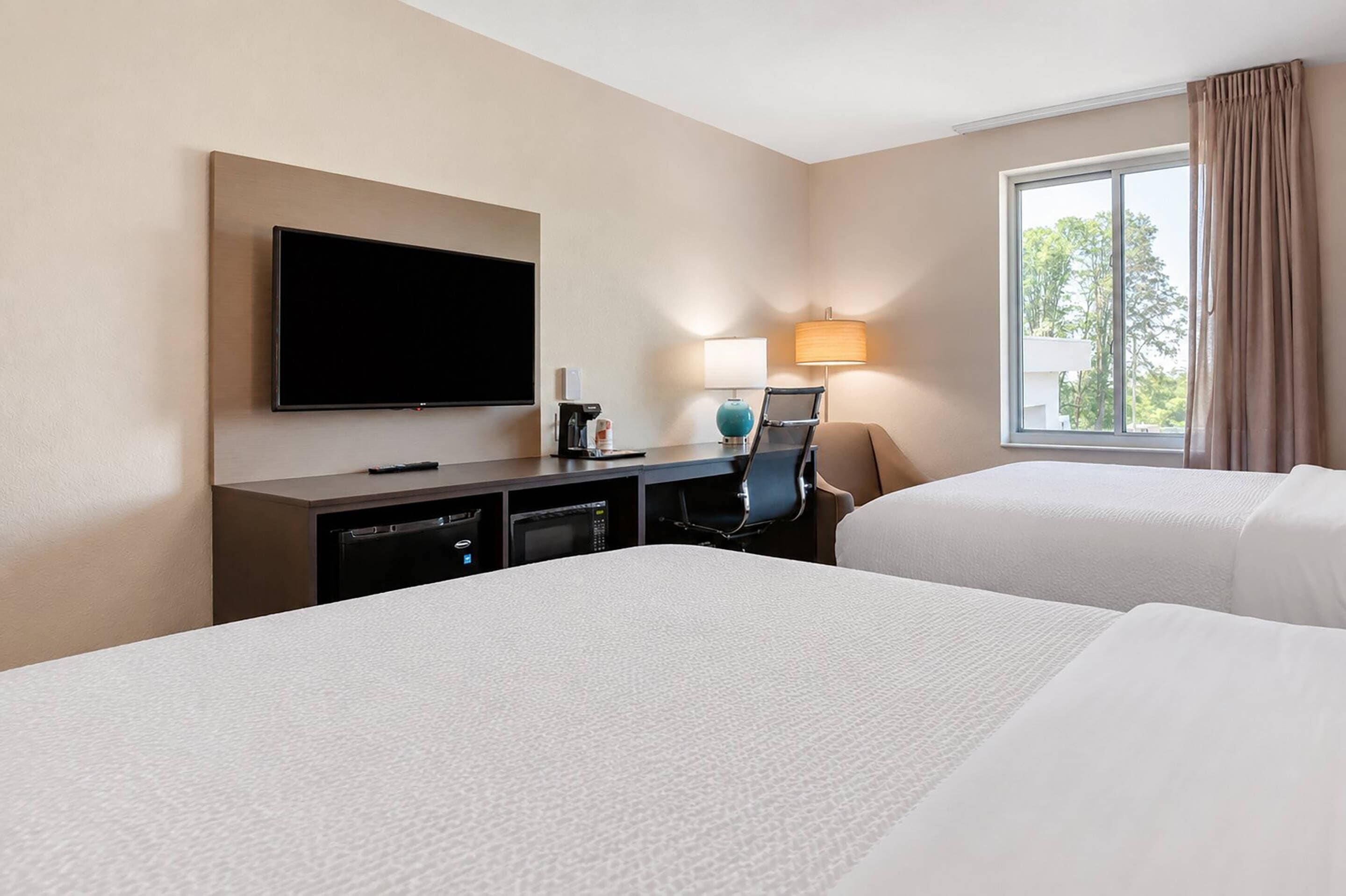 Quarto de hotel com duas camas, televisão, escrivaninha com abajur e janela com cortinas.