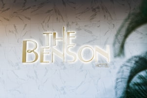 Placa de hotel com o nome 'The Benson Hotel' em letras iluminadas, ao lado de uma parede de mármore.