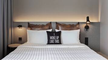 Cama bem arrumada com roupa de cama listrada, travesseiro com a frase 'Acordei assim' e luminária fixada na parede.