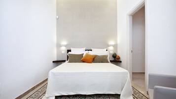 Uma cama bem arrumada com roupa de cama branca e duas almofadas, um piso estampado e uma pequena mesa de cabeceira com um abajur.