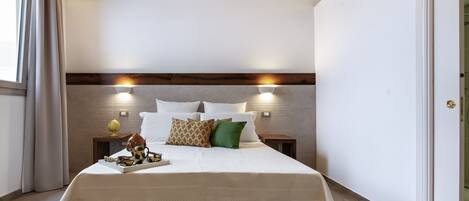 Uma cama bem arrumada com colcha e almofadas brancas, uma mesinha de cabeceira com abajur, e uma janela com cortinas.