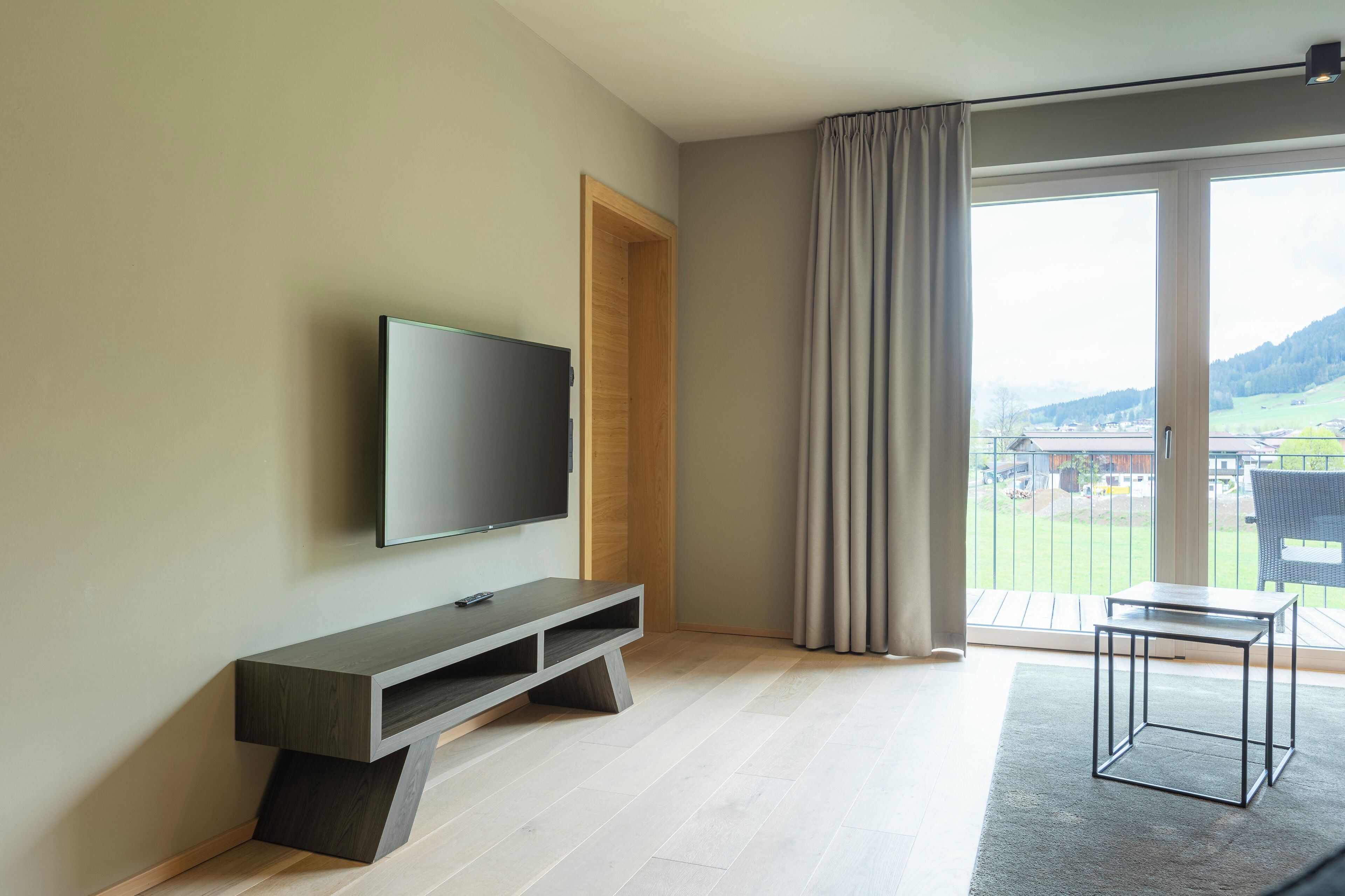 Sala de estar moderna com televisão de tela plana, porta de madeira e vista para uma paisagem verde através de grandes janelas.