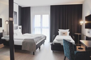 Quarto de hotel com duas camas, uma escrivaninha, uma cadeira, televisão e uma janela grande com cortinas.