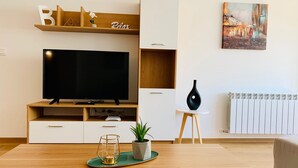 Uma sala de estar moderna com televisão de tela plana, prateleiras de madeira, um armário branco, uma mesinha com uma planta e um radiador.