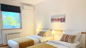 Quarto de hotel com duas camas, um radiador, ar condicionado, janela com vista para árvores e uma obra de arte colorida na parede.