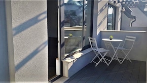Uma varanda com uma cadeira e mesa brancas, uma planta em vaso e vista para um edifício.