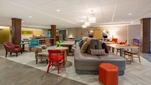 Um lobby moderno de hotel com um sofá, cadeiras vermelhas, uma área de refeições e uma cozinha ao fundo.