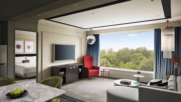 Quarto de hotel moderno com uma janela grande, televisão de tela plana, poltrona vermelha, sofá, mesa de jantar com maçãs e uma pequena mesinha de apoio.