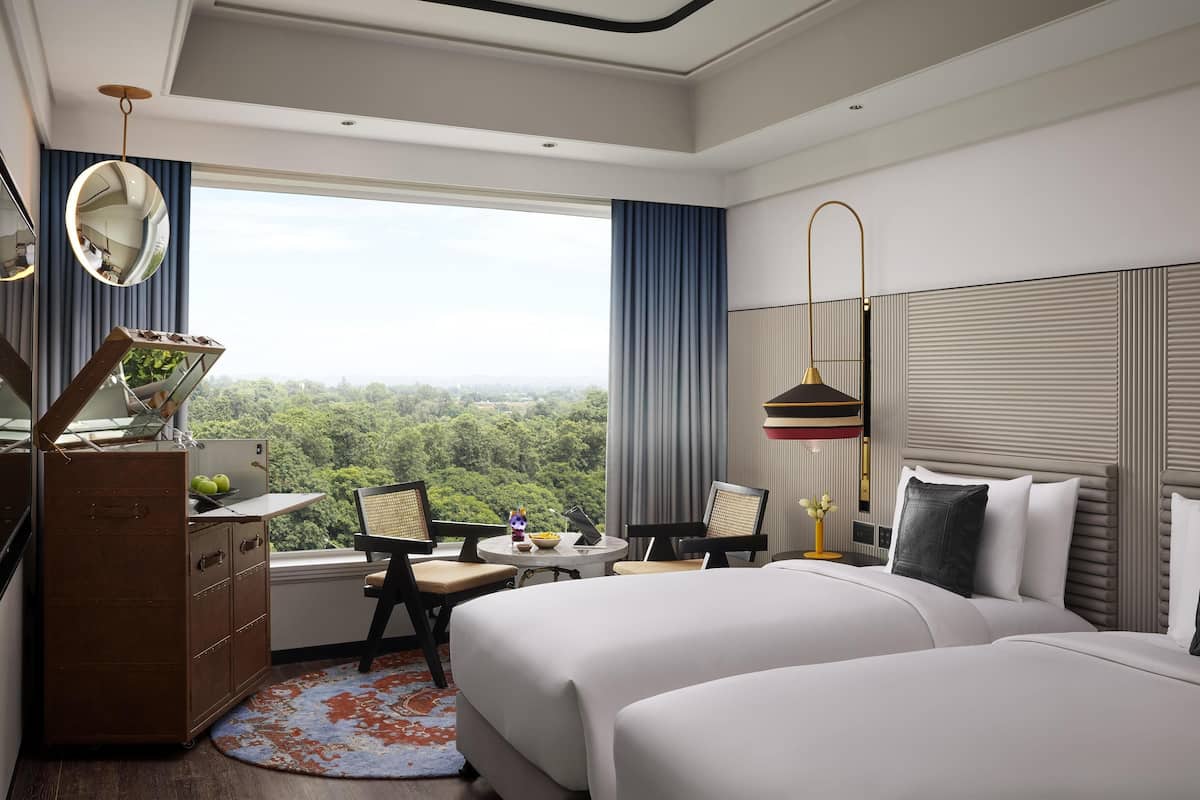 Quarto de hotel com duas camas, uma escrivaninha, uma cadeira, uma mesinha, um espelho e vista para o exterior.