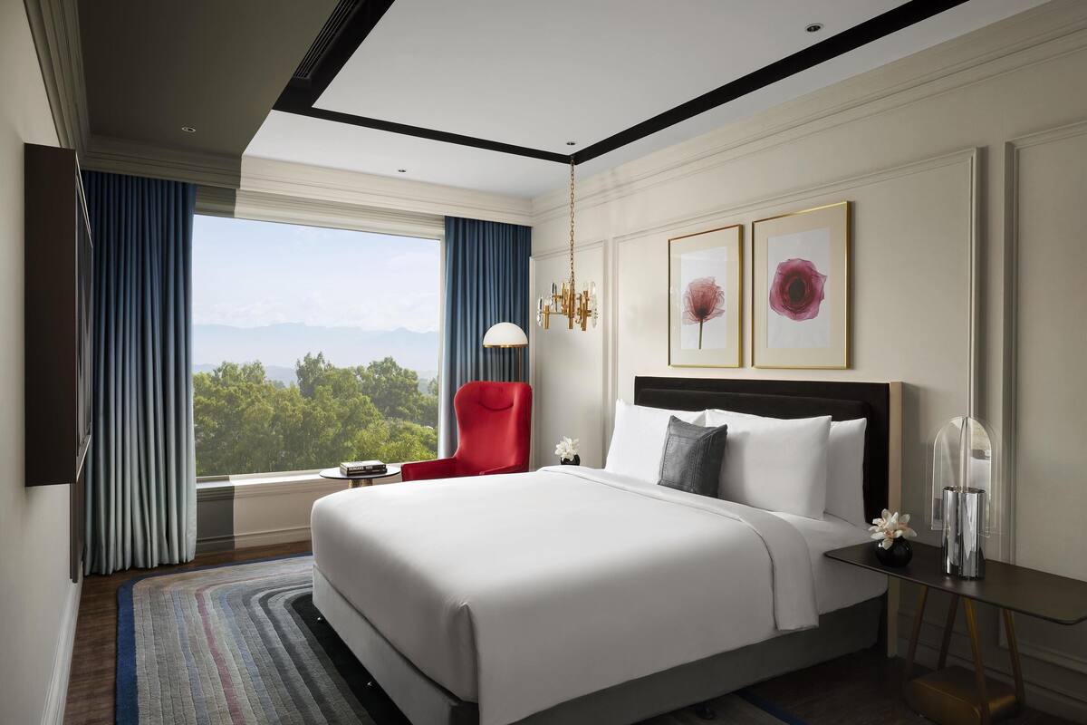 Quarto de hotel moderno com uma cama grande, uma cadeira vermelha, uma mesa de cabeceira e vista para o exterior.