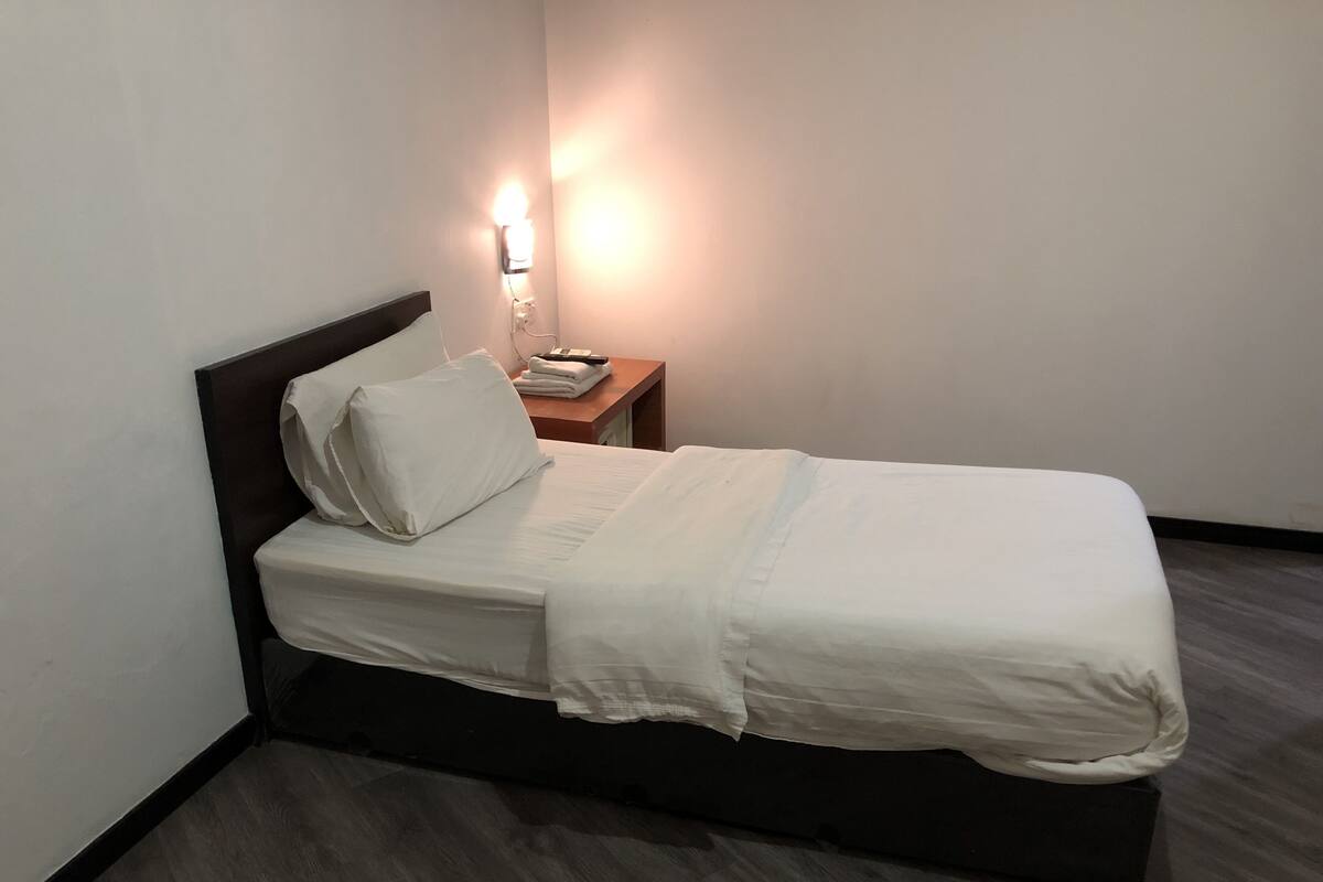 Cama bem arrumada com edredom branco e travesseiros, mesa de cabeceira com abajur e cabeceira de madeira escura.