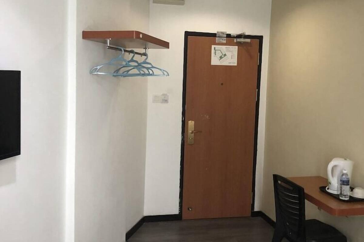 Quarto de hotel com uma cama, uma escrivaninha com cadeira, porta de madeira e um cabideiro fixado na parede.