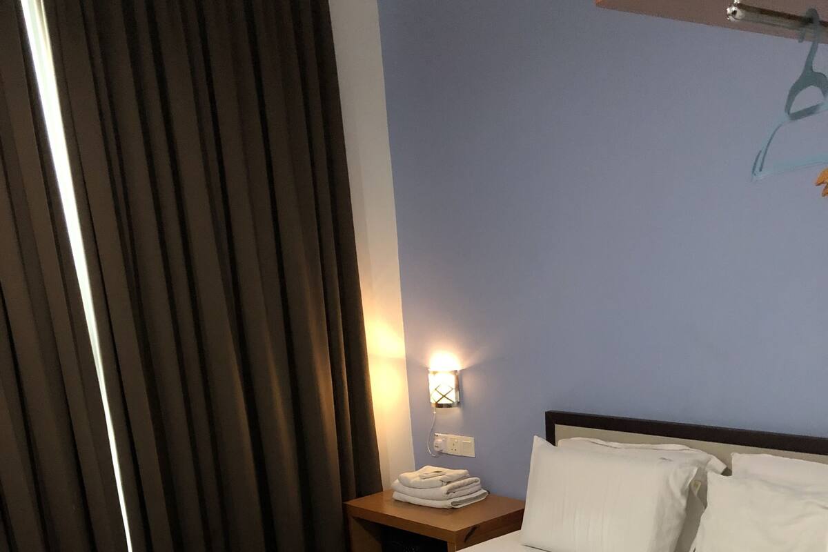 Cama bem arrumada com lençóis brancos, mesa de cabeceira de madeira com abajur e um cabideiro.