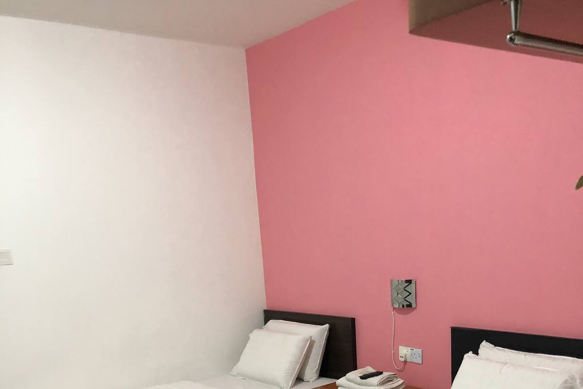 Quarto de hotel com duas camas de solteiro, uma mesinha de cabeceira de madeira com abajur e uma parede rosa.