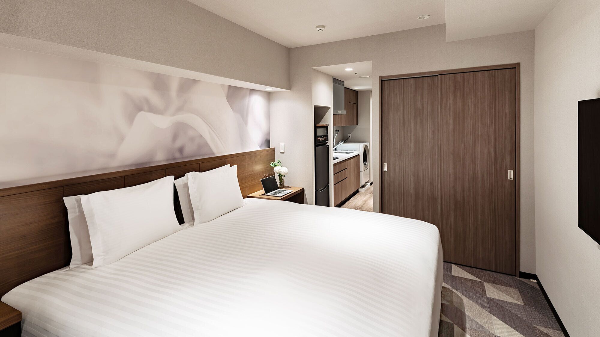 Quarto de hotel com uma cama grande, uma mesa de cabeceira, uma escrivaninha com um laptop, uma televisão na parede e um banheiro visível por uma porta aberta.