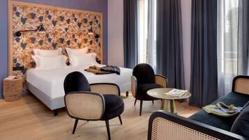 Quarto de hotel com cama, duas cadeiras, uma mesa pequena e uma janela grande com cortinas.