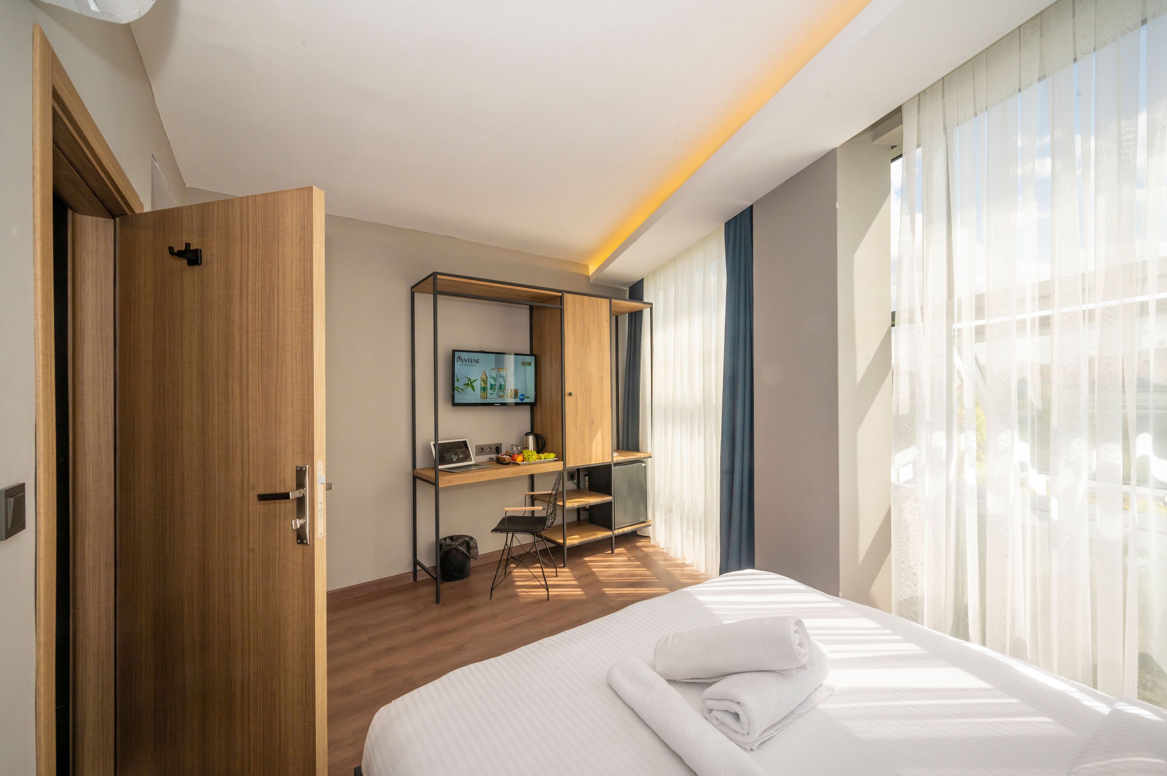 Quarto de hotel moderno com porta de madeira, cama com roupa branca, escrivaninha com um laptop, pequena área de estar e uma janela grande com cortinas leves.