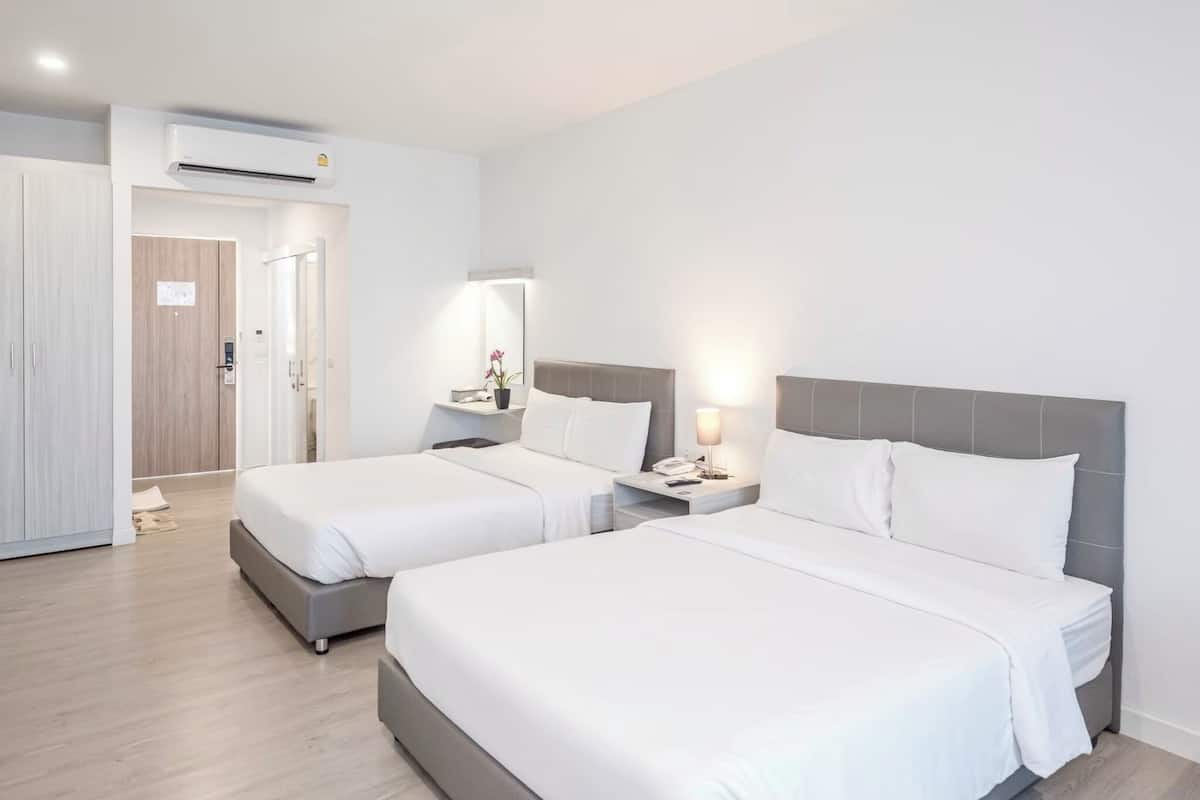 Quarto de hotel com duas camas, uma mesa de cabeceira com abajur, um aparelho de ar condicionado e uma porta que dá para outro cômodo.