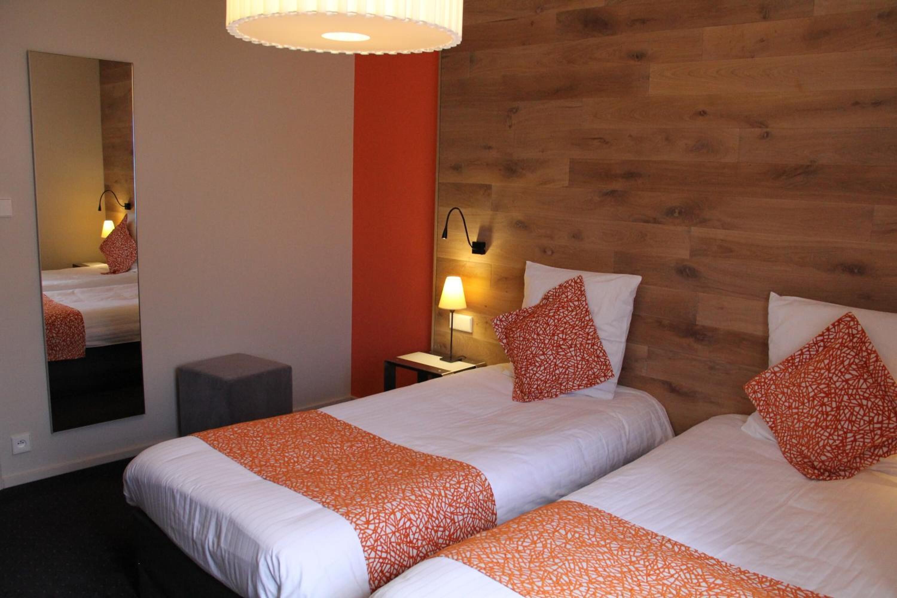 Quarto de hotel com duas camas, um espelho, uma mesa de cabeceira com abajur e uma parede de madeira.