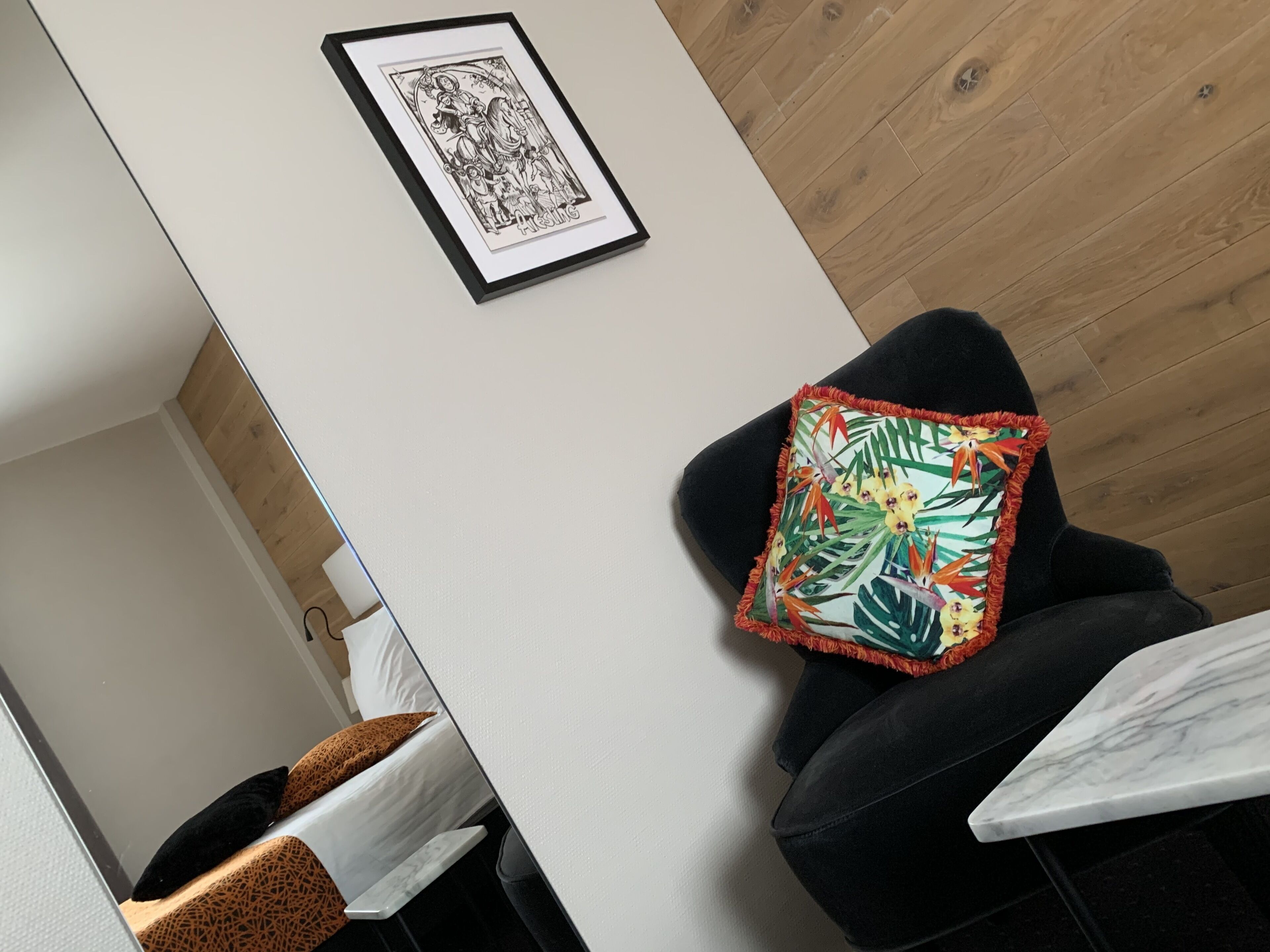 Um quarto com piso de madeira, uma cadeira preta com almofada colorida, uma parede branca com um quadro emoldurado e uma cama com almofadas estampadas.