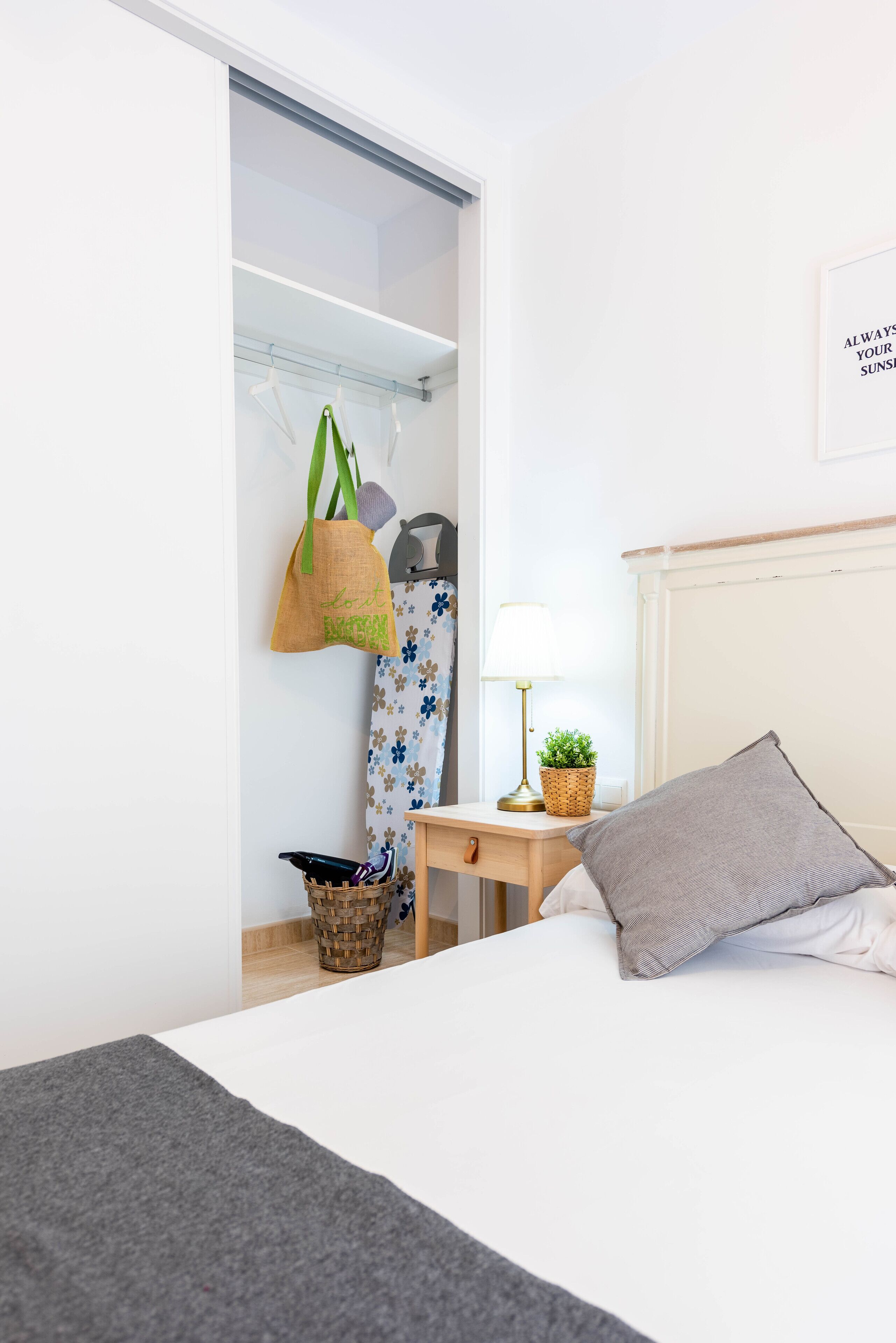 Quarto com cama, mesa de cabeceira com abajur, planta em vaso, cesta e bolsa pendurada.