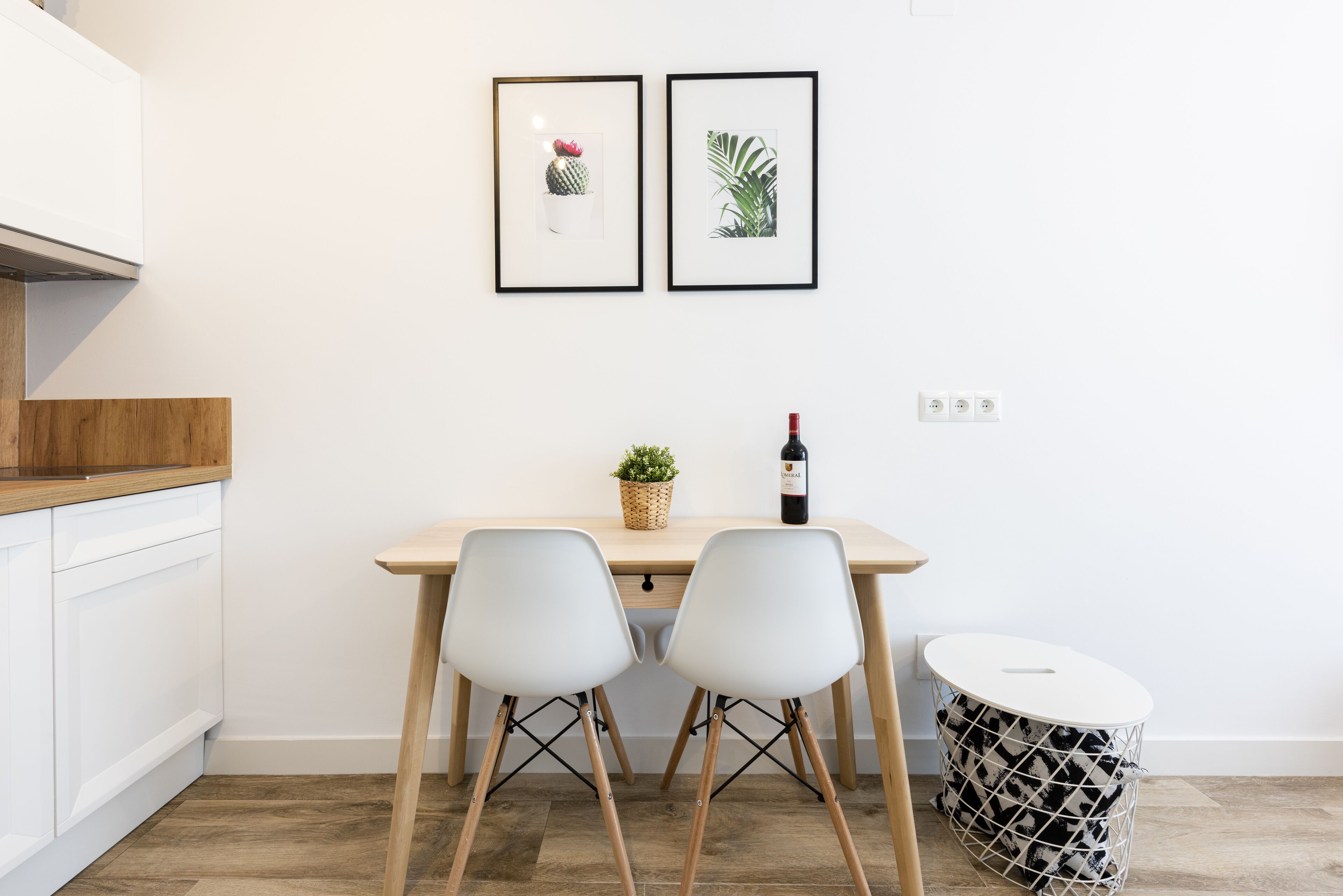 Cozinha moderna com armários brancos, bancada de madeira, duas cadeiras brancas, uma pequena mesa redonda com uma garrafa de vinho e duas fotos emolduradas na parede.