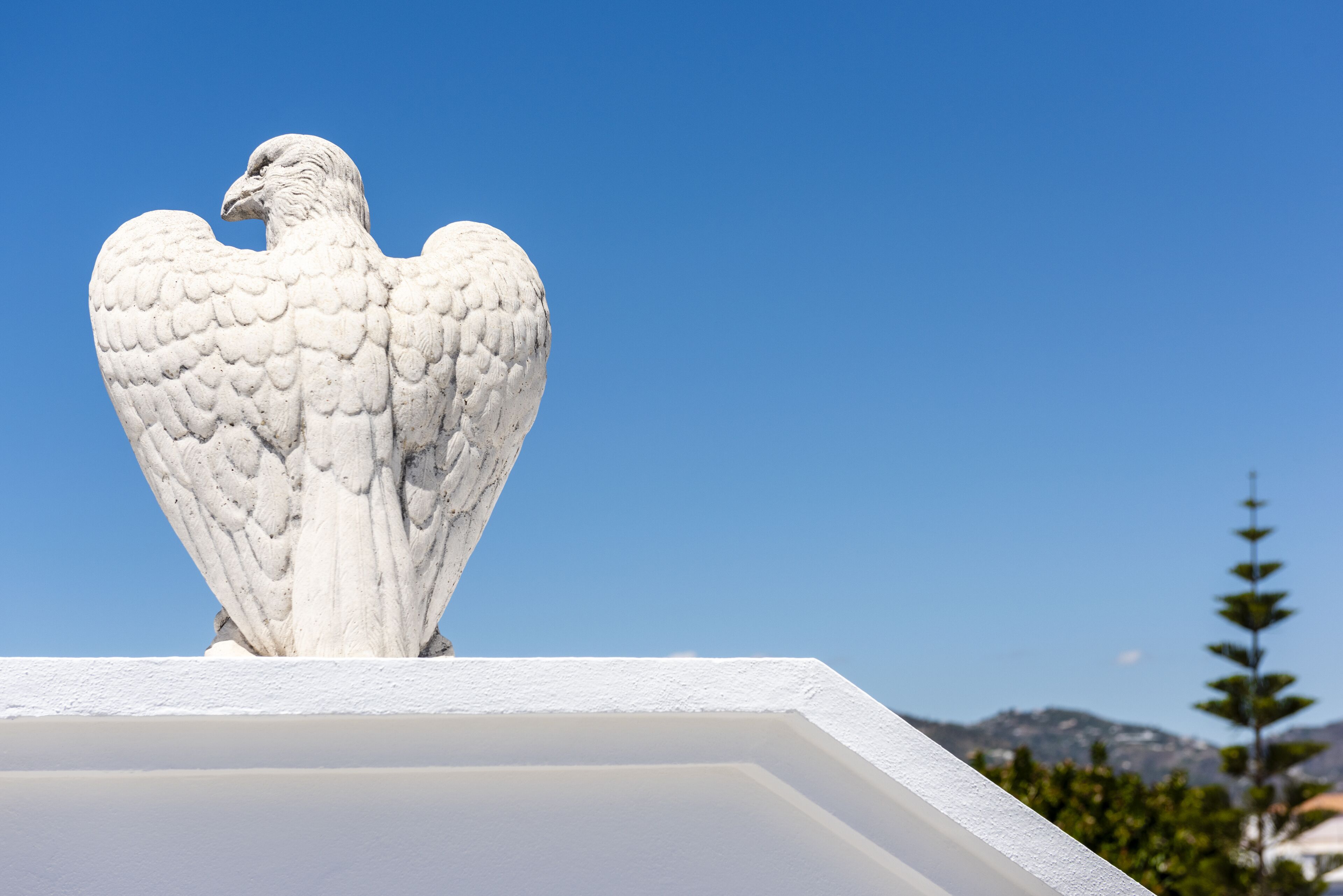 Uma estátua branca de águia sobre um pedestal branco, com um céu azul límpido e montanhas distantes ao fundo.
