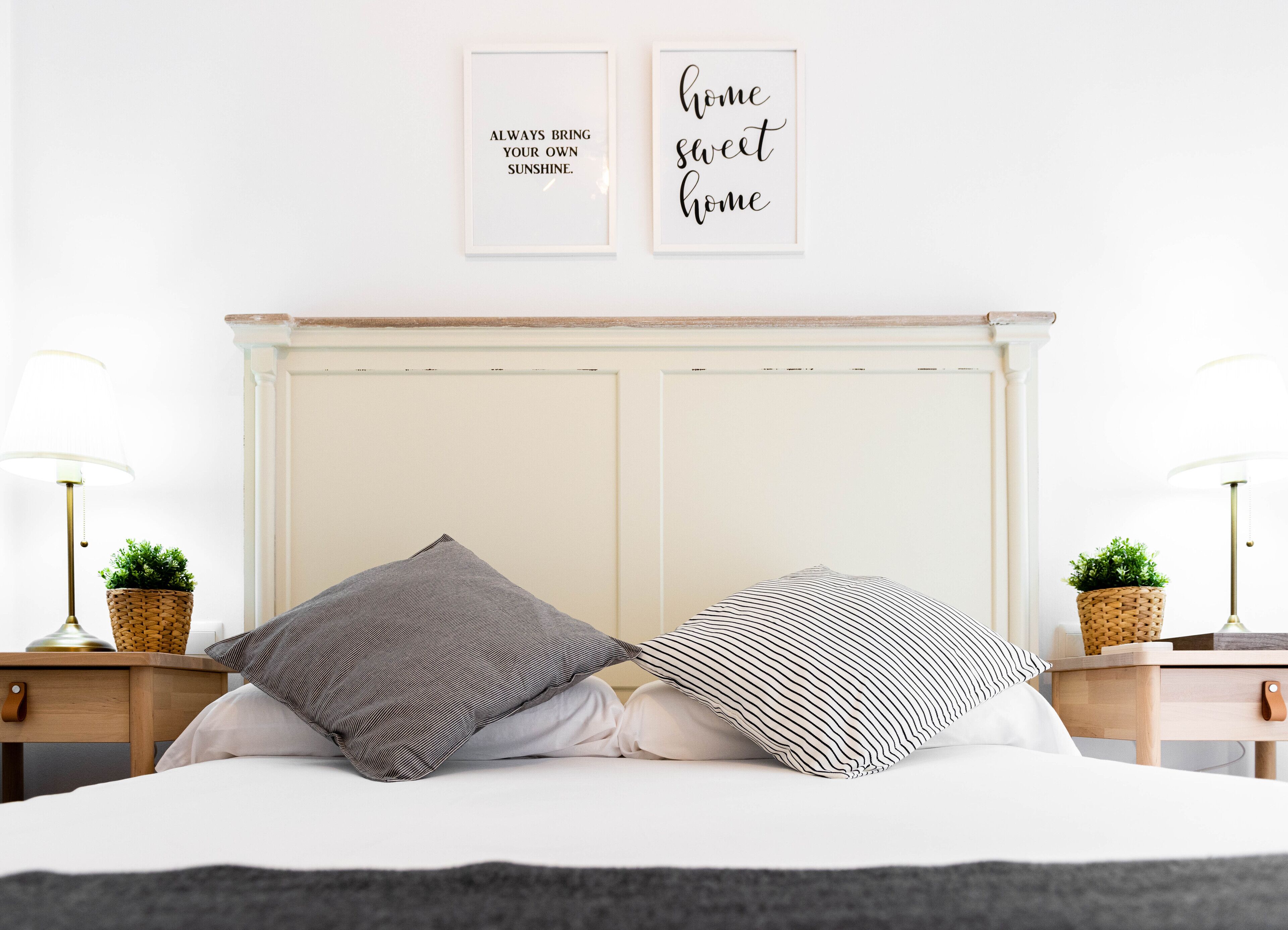 Uma cama bem arrumada com duas almofadas, uma mesinha de cabeceira com abajur, e duas frases inspiradoras emolduradas na parede.