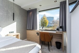 Quarto de hotel moderno com uma cama grande, uma secretária de madeira, uma cadeira, uma cafeteira e vista para a paisagem urbana pela janela.