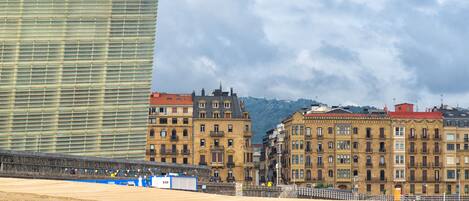 Paisagem costeira urbana com mistura de edifícios modernos e históricos, uma praia arenosa e um céu nublado.