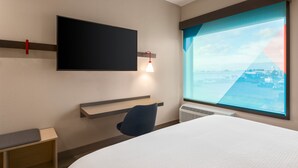 Quarto de hotel moderno com cama, escrivaninha, cadeira, televisão de tela plana na parede e uma grande janela com vista para a cidade.
