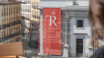 Uma pessoa sentada à mesa com uma bebida e livros, com vista para um horizonte urbano repleto de edifícios históricos e um banner vermelho com texto.