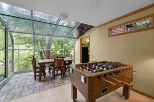 Quarto com mesa de pebolim, mesa de jantar com cadeiras e amplas janelas com vista para árvores.