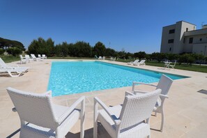 Piscina com cadeiras de descanso brancas e um edifício ao fundo.