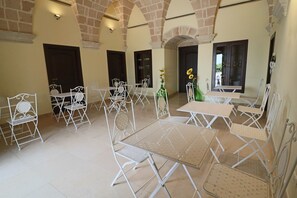 Amplo espaço interno com diversas mesas e cadeiras de metal branco dispostas em semicírculo, cercado por arcos de tijolo e grandes janelas.