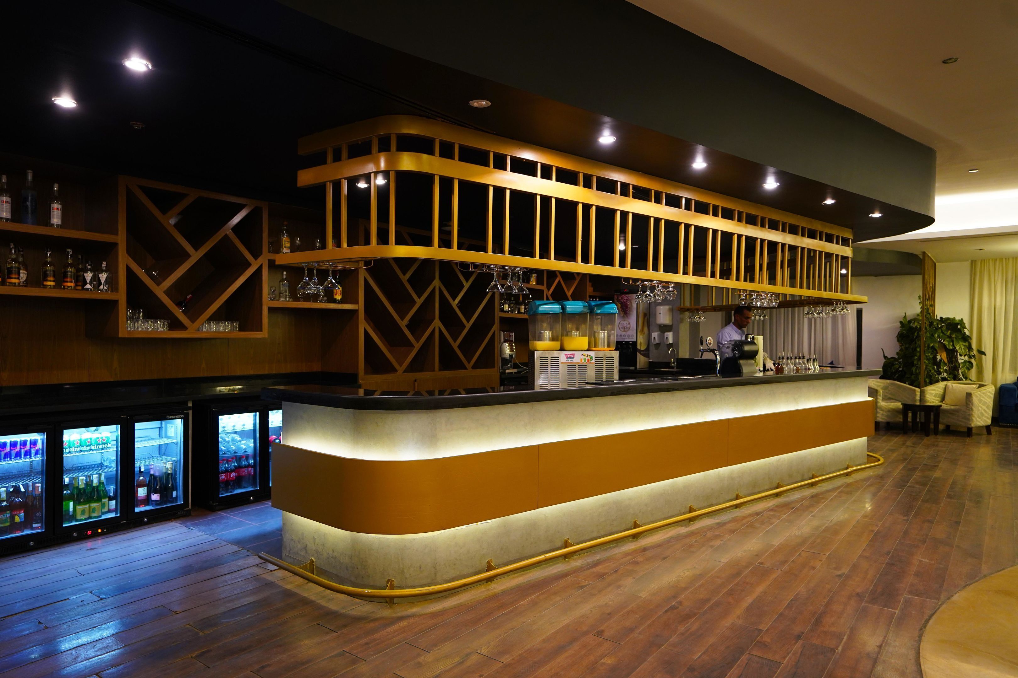Uma área de bar com balcão curvo, prateleiras com garrafas e um espaço de estar com uma planta.