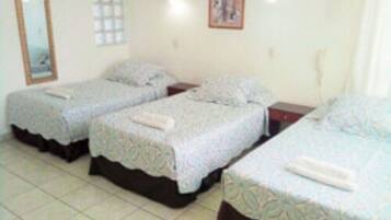 Quarto com três camas de solteiro, cada uma com travesseiro e cobertor. Há um espelho na parede e uma mesinha de cabeceira ao lado de uma cama.
