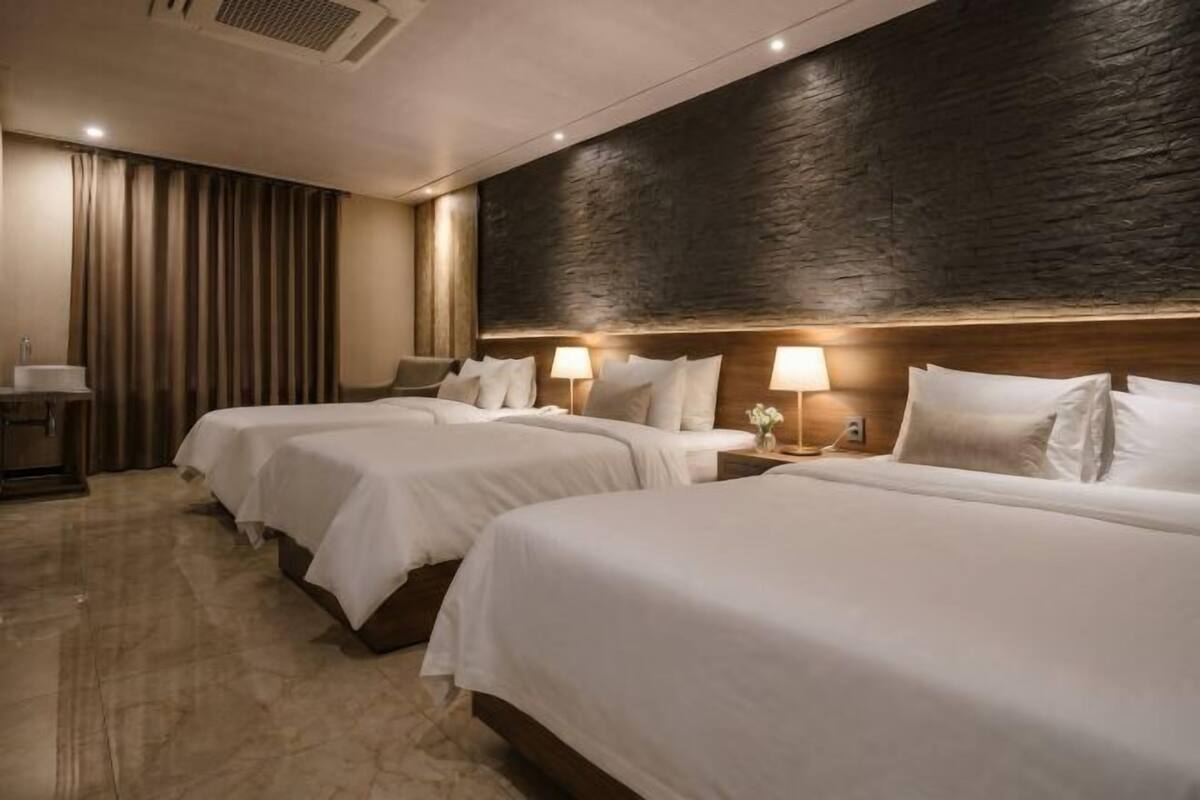 Quarto de hotel com três camas, cabeceira de madeira, mesa de cabeceira com abajur e uma janela grande com cortinas.