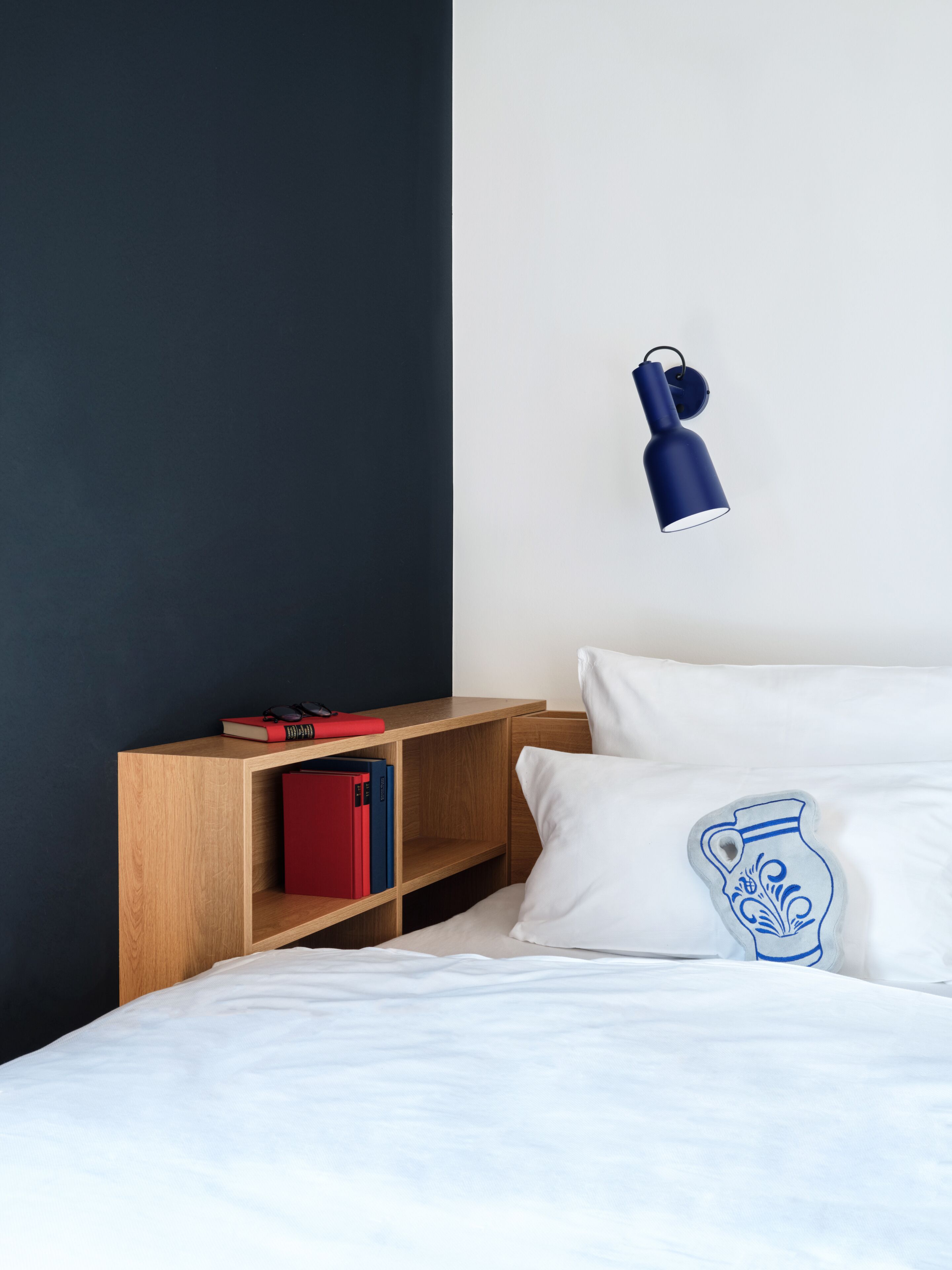 Quarto com mesa de cabeceira de madeira, abajur azul, travesseiro branco com desenho azul bordado e um livro vermelho.