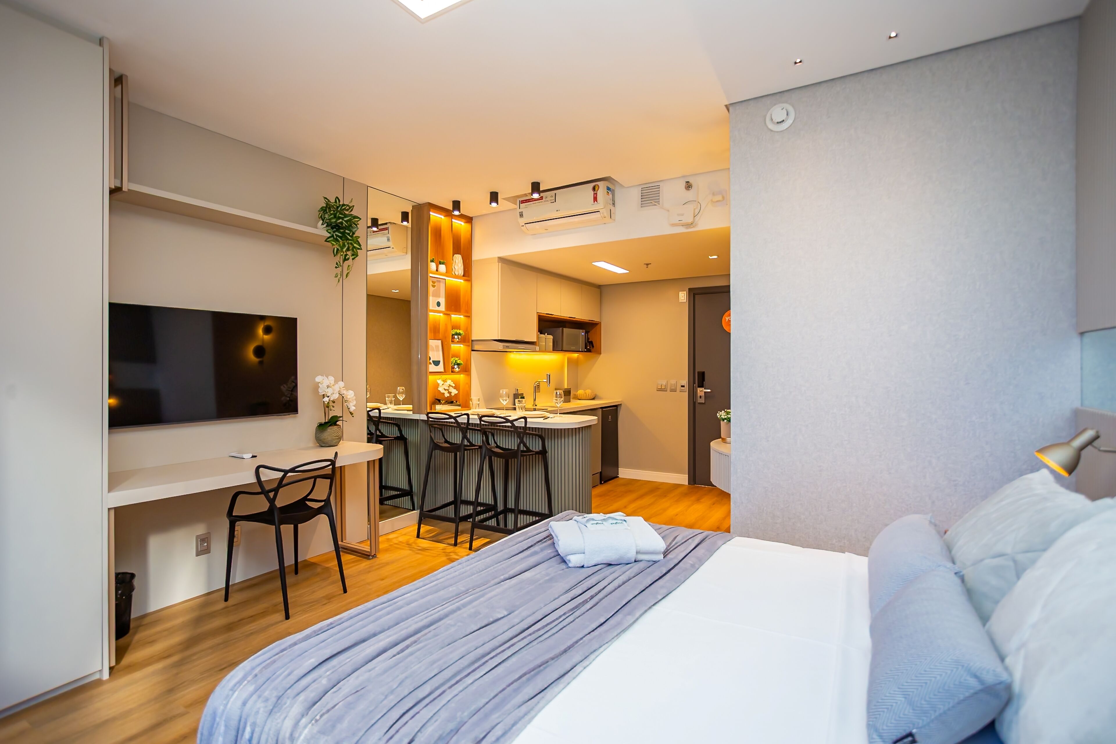 Quarto de hotel moderno com uma cama grande, uma escrivaninha com cadeira, uma pequena cozinha e uma televisão.