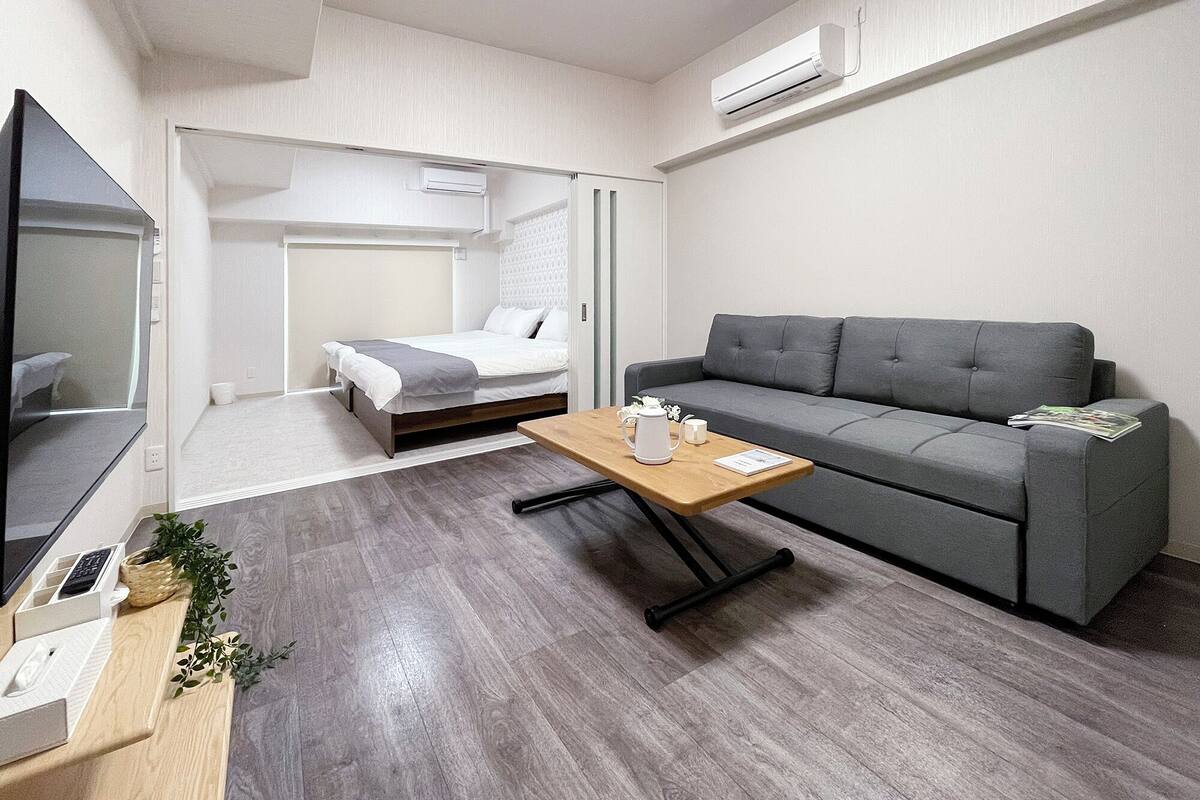 Quarto de hotel moderno e compacto, com cama, sofá e uma pequena mesa com chaleira e chávenas.