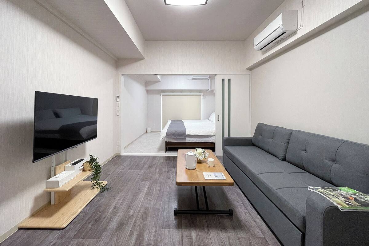 Quarto de hotel moderno com televisão de tela plana, sofá, mesa de centro de madeira e cama com vista para o corredor.